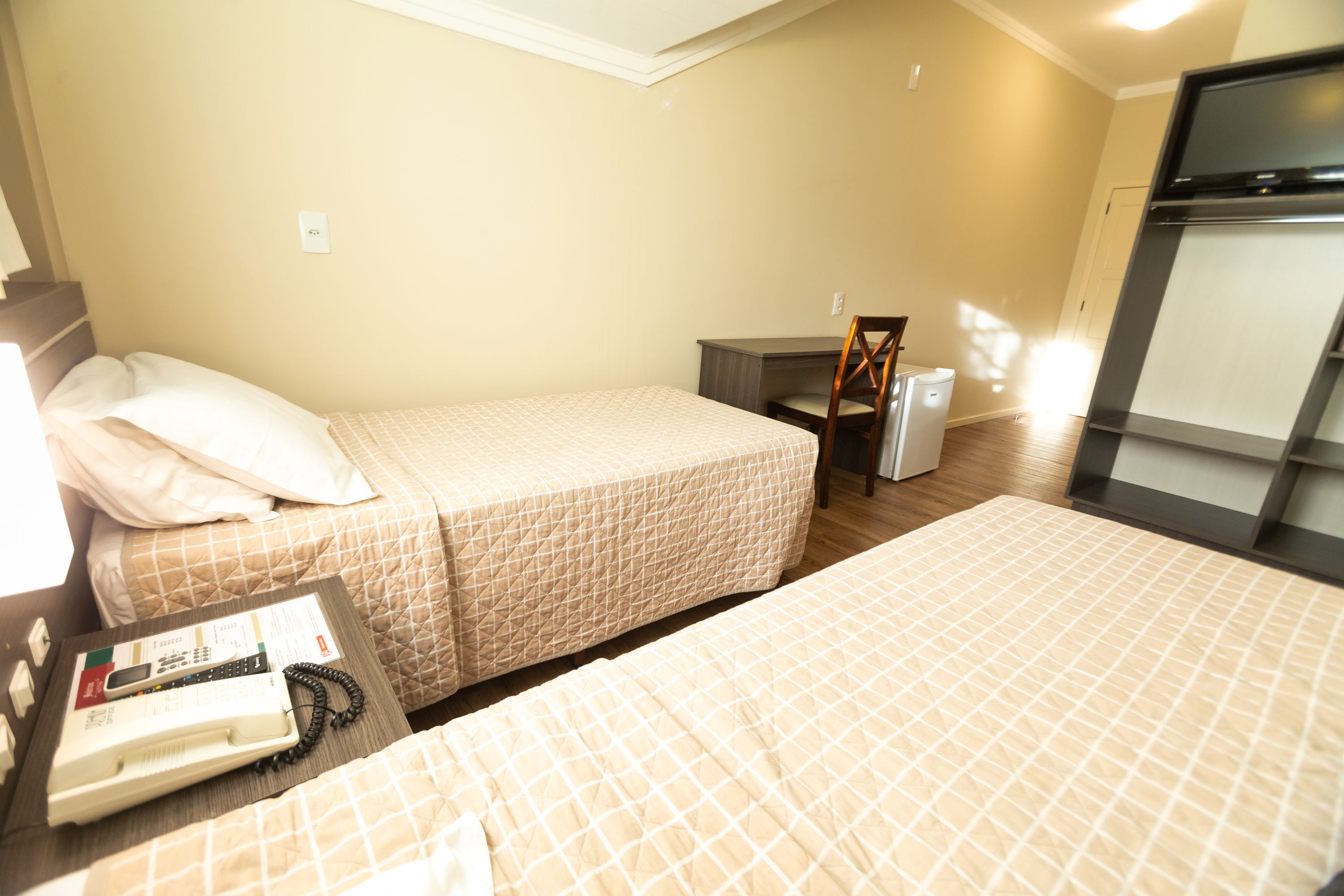 Quarto de hotel com duas camas, uma escrivaninha, uma cadeira, um refrigerador e um telefone.