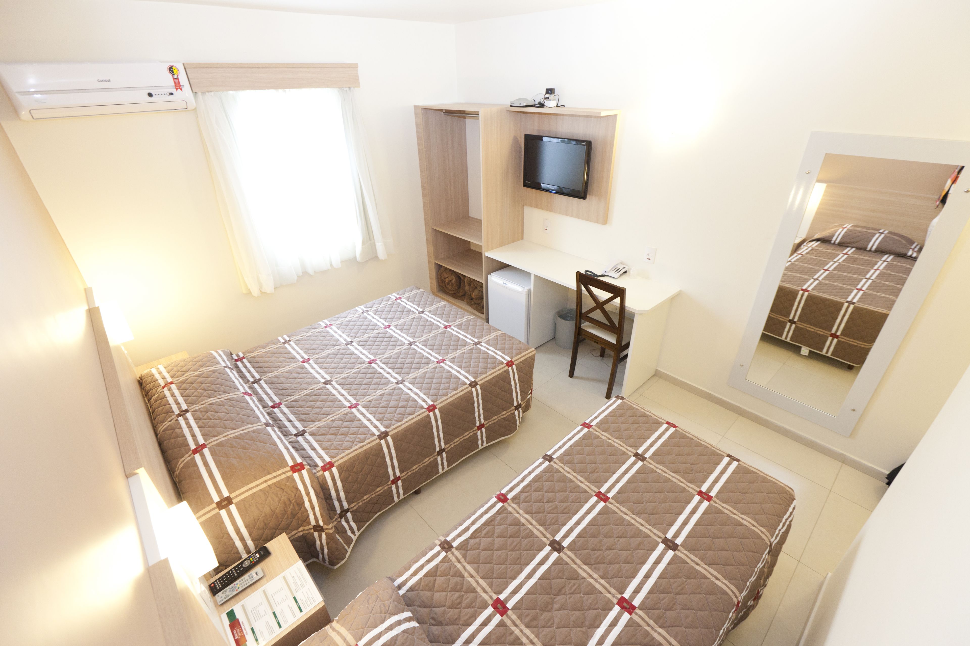 Quarto de hotel compacto com duas camas, uma mesa, televisão e espelho.