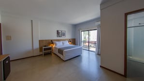 Quarto de hotel moderno com uma cama grande, mesa de cabeceira com abajur, prateleira pequena e janela com vista para edifícios.