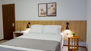 Quarto arrumado com cama, mesas de cabeceira, uma pequena mesa redonda com uma planta e duas molduras na parede.