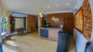 Uma área de recepção de hotel com um balcão de madeira, um espaço de estar com cadeiras e uma grande janela com plantas.