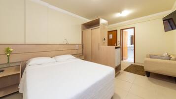 Quarto de hotel com uma cama grande, um guarda-roupa de madeira, uma pequena mesa de trabalho com uma luminária e um sofá.