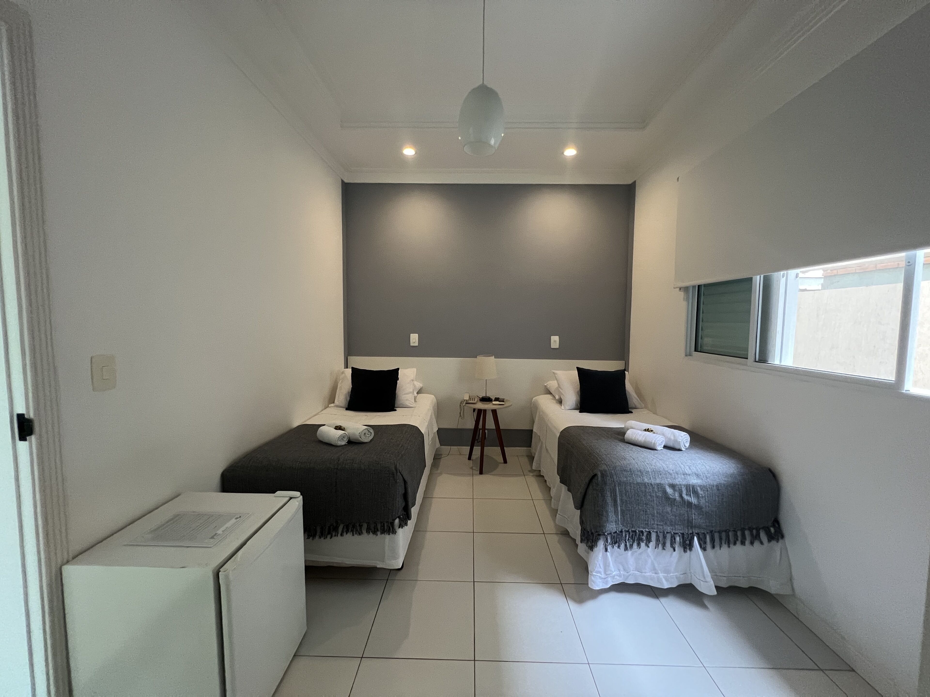 Quarto pequeno e limpo com duas camas, uma mesinha e uma janela.