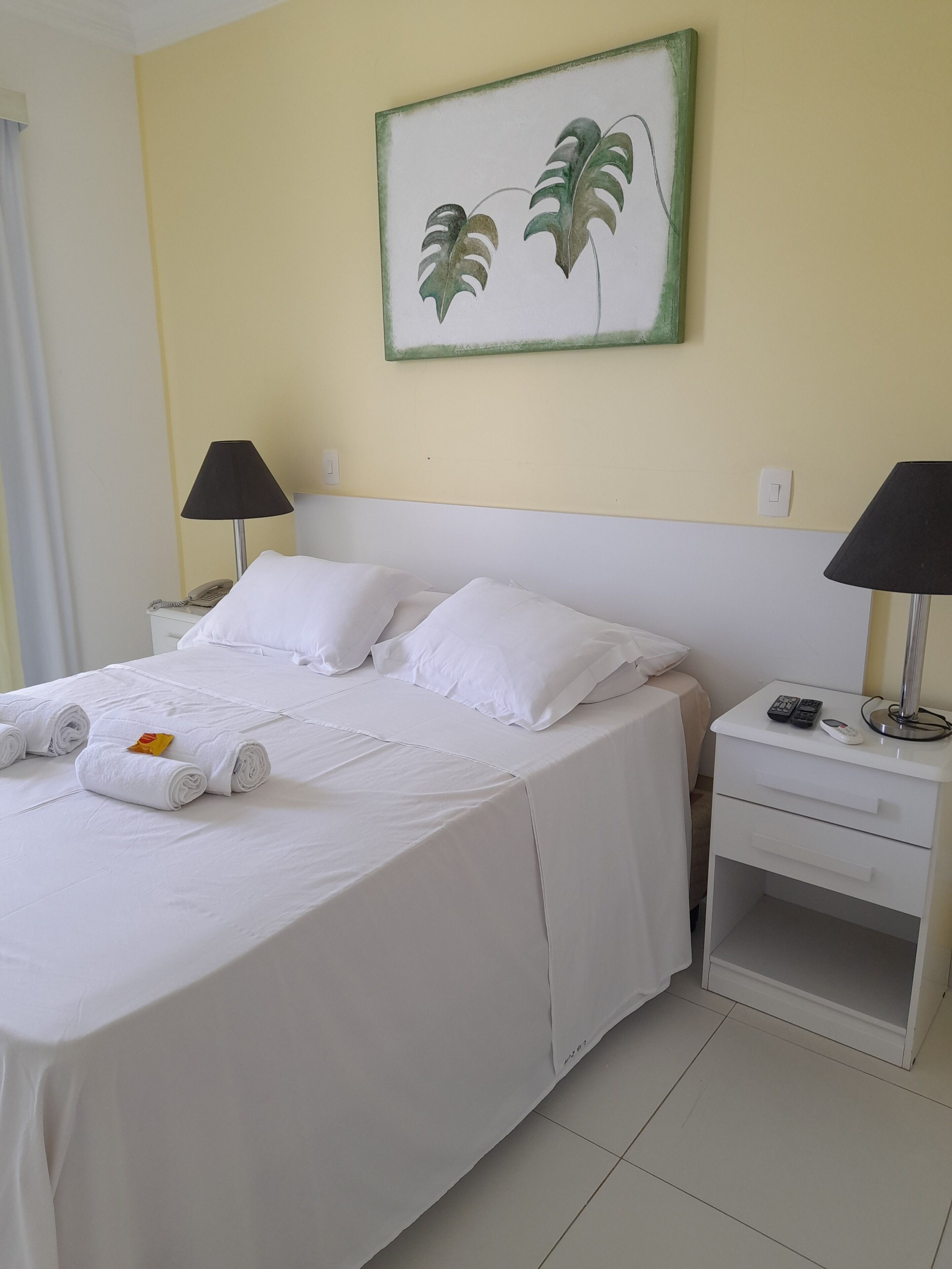Cama bem arrumada com lençóis brancos e toalhas enroladas, mesa de cabeceira com abajur e um quadro na parede com folhas.