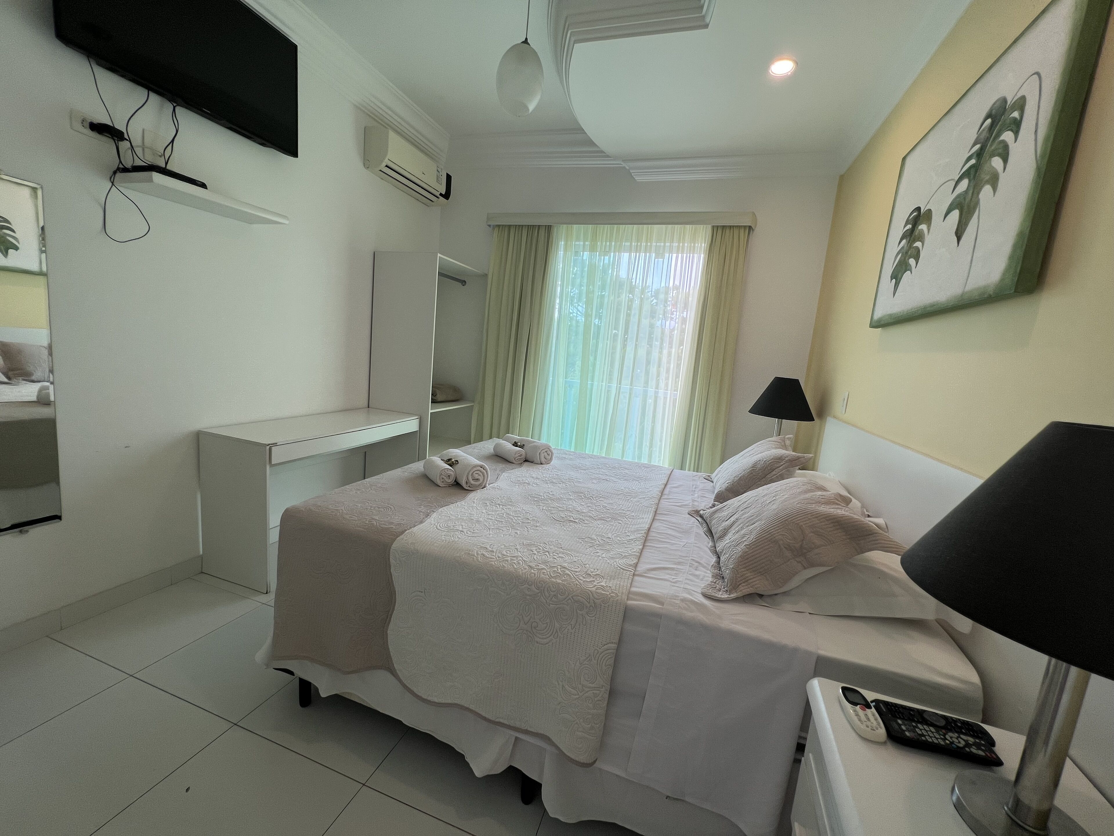 Quarto de hotel com cama, mesa de cabeceira, luminária e escrivaninha com cadeira.