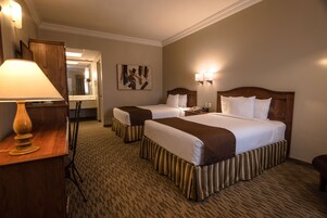 Quarto de hotel com duas camas, uma escrivaninha, uma luminária e um espelho.