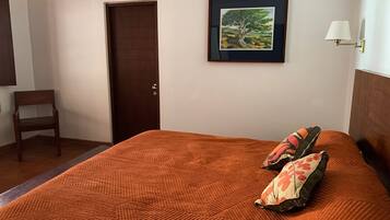 Um quarto com uma cama grande, uma porta de madeira, um quadro emoldurado, uma cadeira e um abajur fixado na parede.