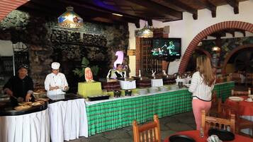 Um restaurante com parede de pedra, mesa de buffet e tela de televisão exibindo uma banda.