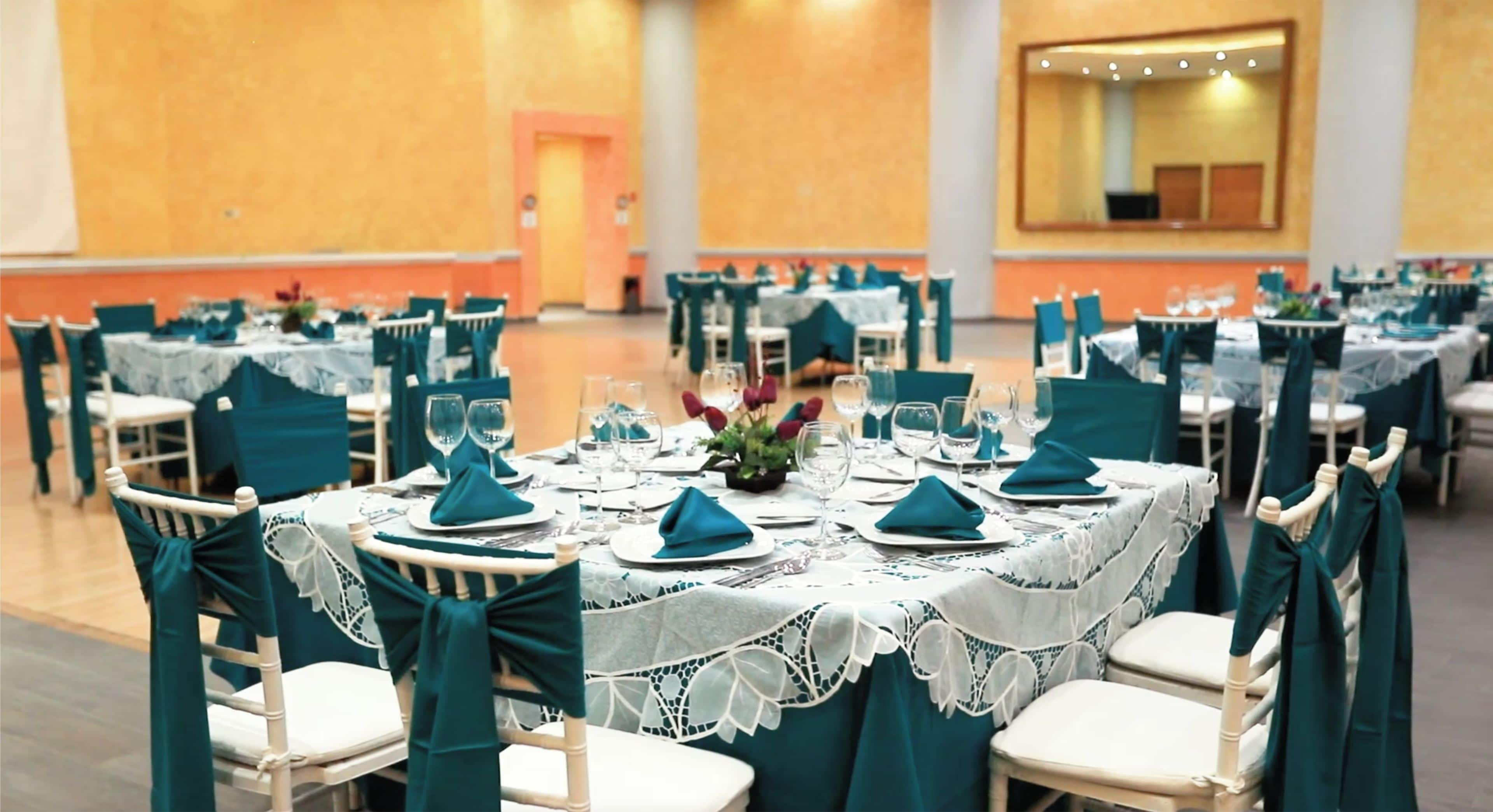 Uma sala de banquetes com mesas redondas preparadas para refeições, cada uma com toalhas de renda branca e capas de cadeira verde-água.
