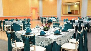 Uma sala de banquetes com mesas redondas preparadas para refeições, cada uma com toalhas de renda branca e capas de cadeira verde-água.