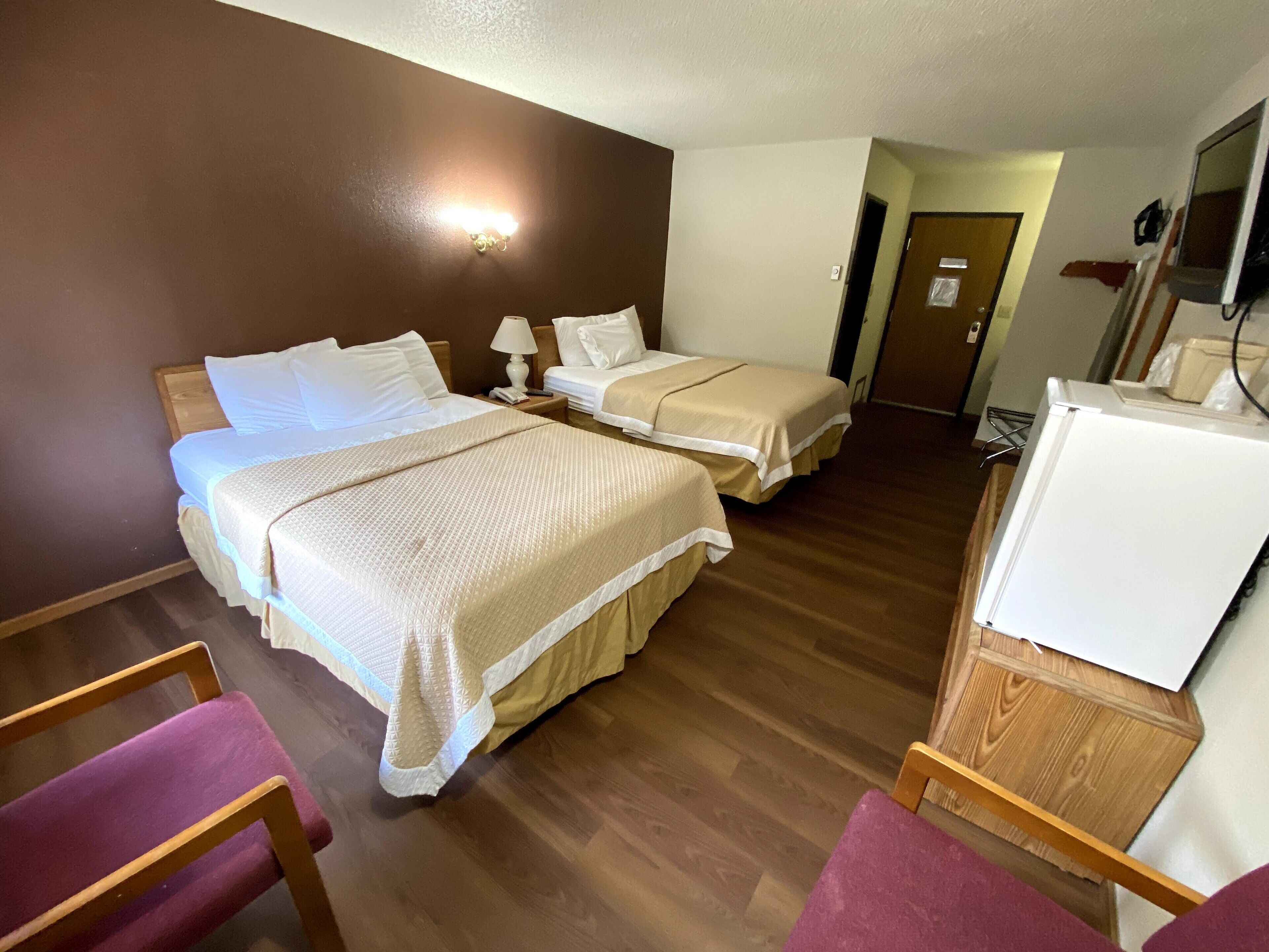 Quarto de hotel com duas camas, piso de madeira, escrivaninha com cadeira e um refrigerador.