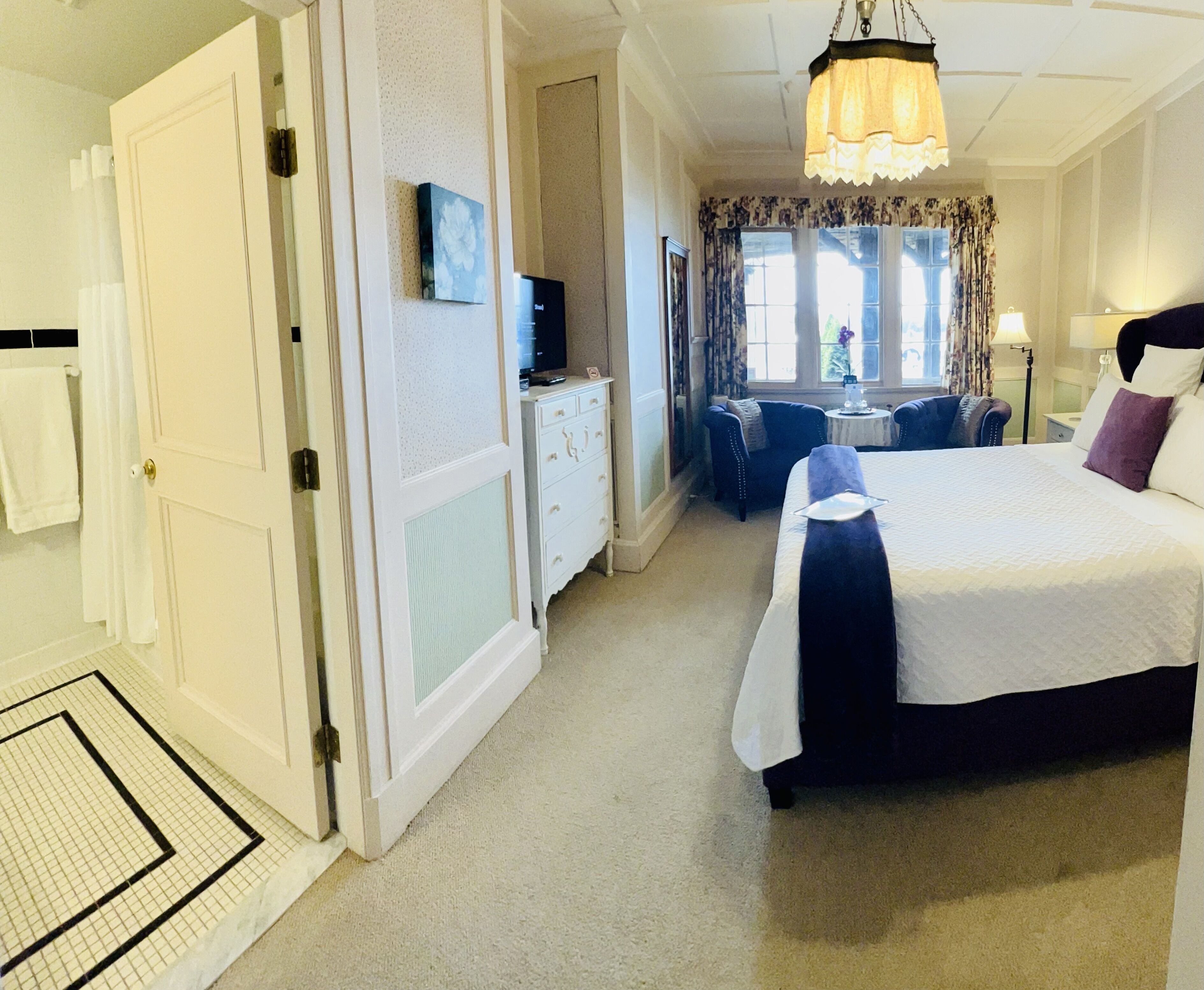Quarto de hotel com cama, escrivaninha, cadeira, televisão e janela com cortinas.