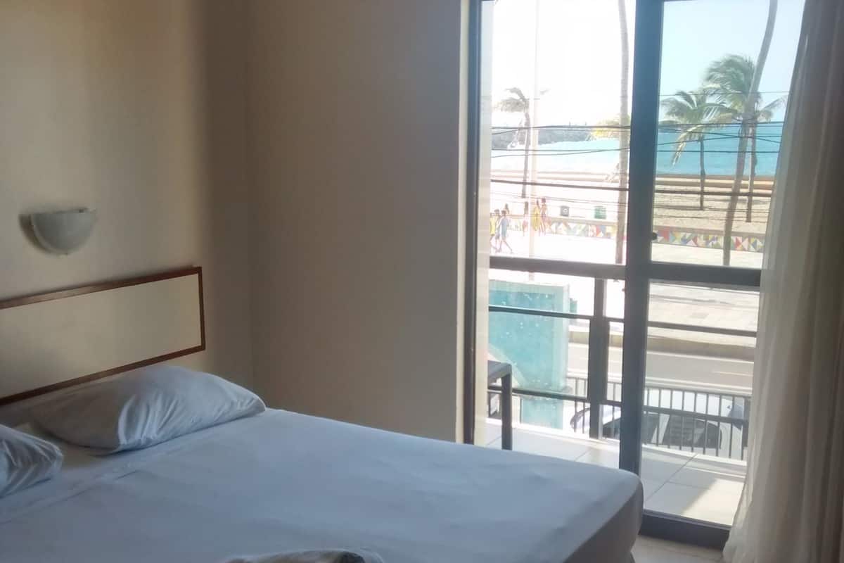 Quarto de hotel com cama, vista para a praia e varanda com guarda-corpo.