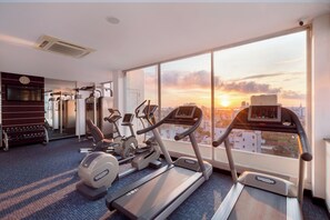 Uma academia com esteiras, elípticos e equipamentos de musculação, com vista para a cidade através de amplas janelas.