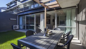 Uma área moderna de refeições ao ar livre, com mesa e cadeiras, guarda-corpo de vidro e vista do interior através de grandes portas deslizantes.