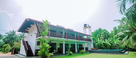 Um edifício de dois andares com varanda, cercado por vegetação exuberante e uma piscina.