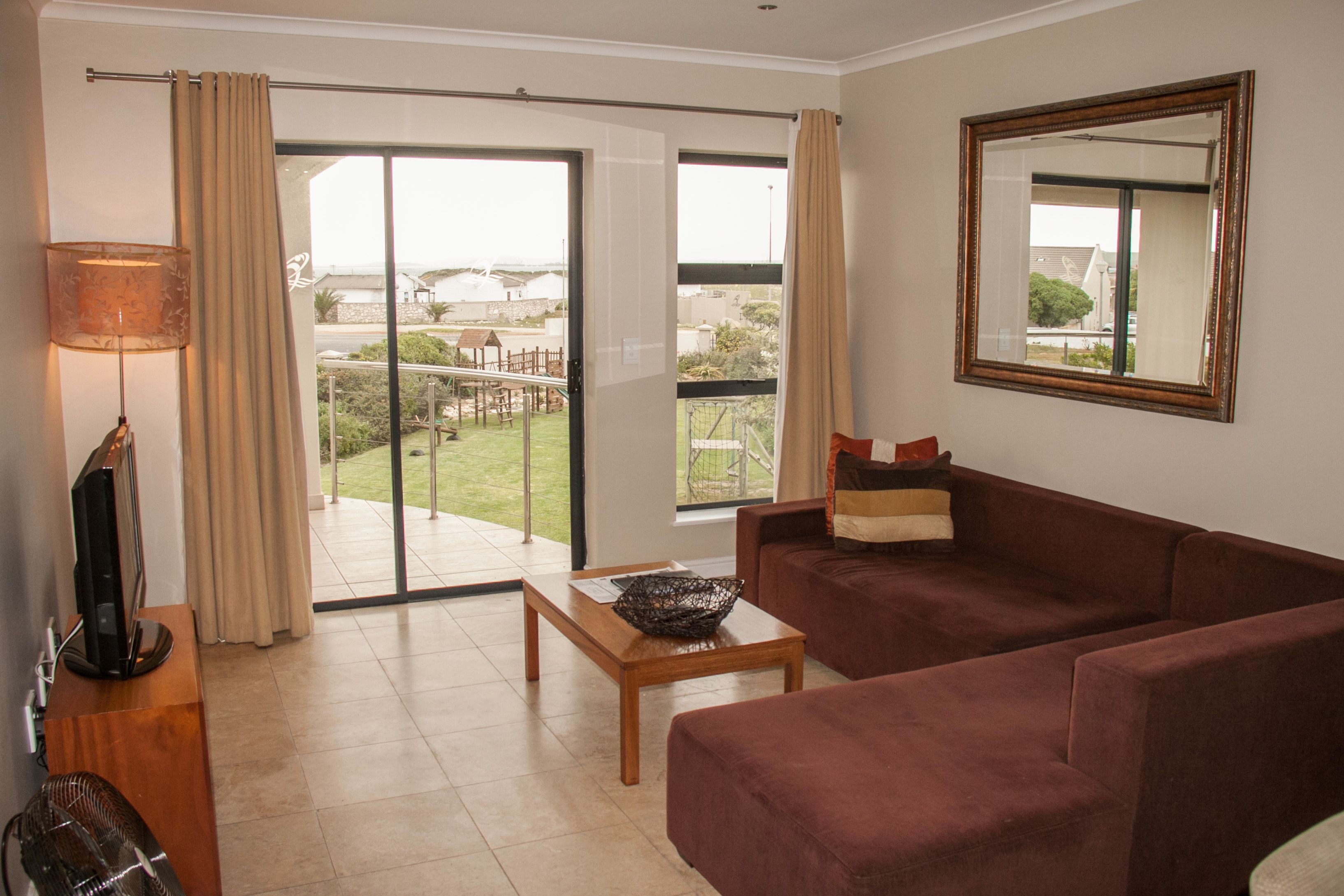 Sala de estar com sofá marrom, mesa de centro de madeira, televisão em suporte de madeira e uma grande janela com cortinas.