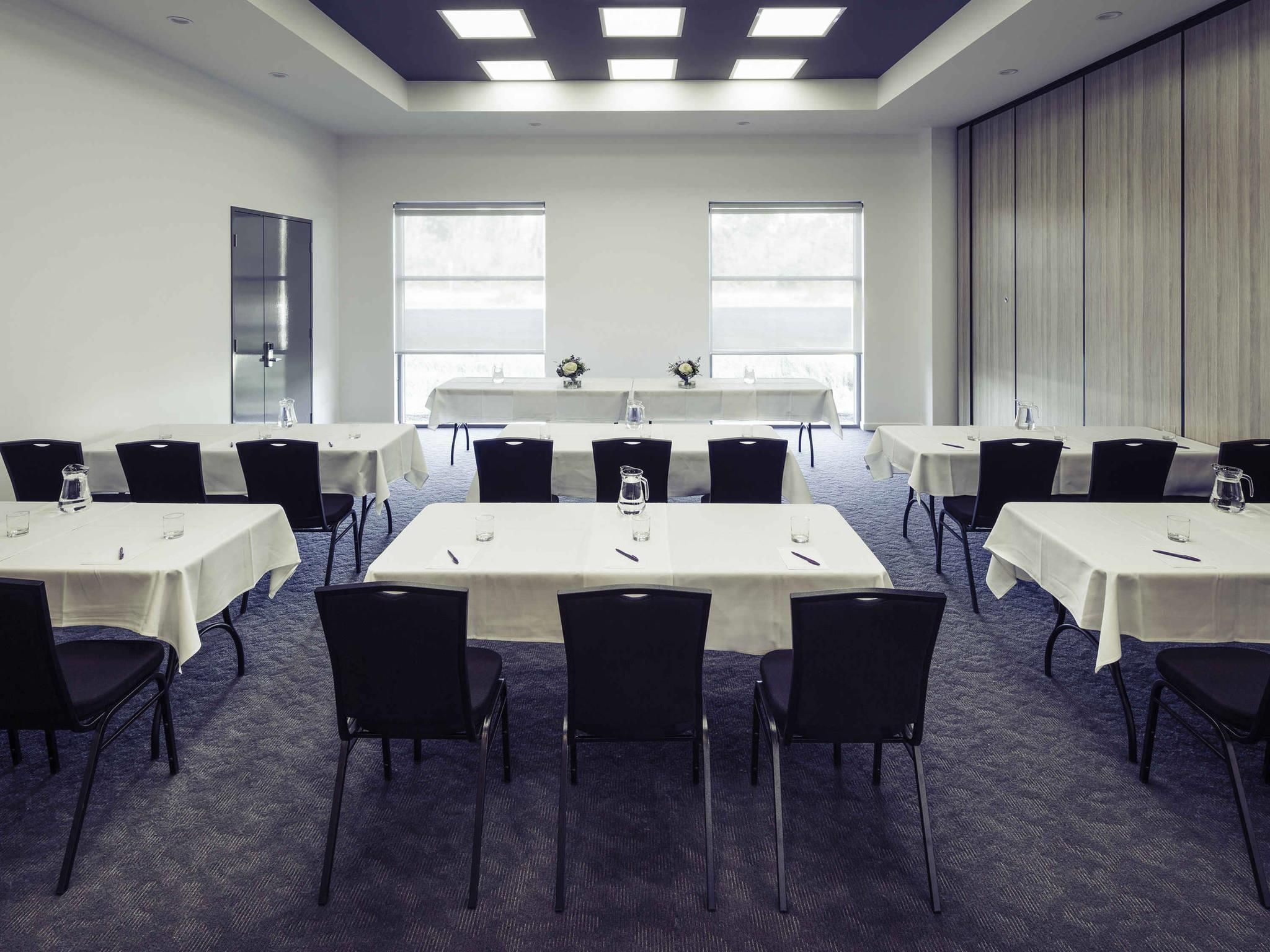 Sala de conferências com várias mesas e cadeiras, janelas amplas e design moderno no teto.
