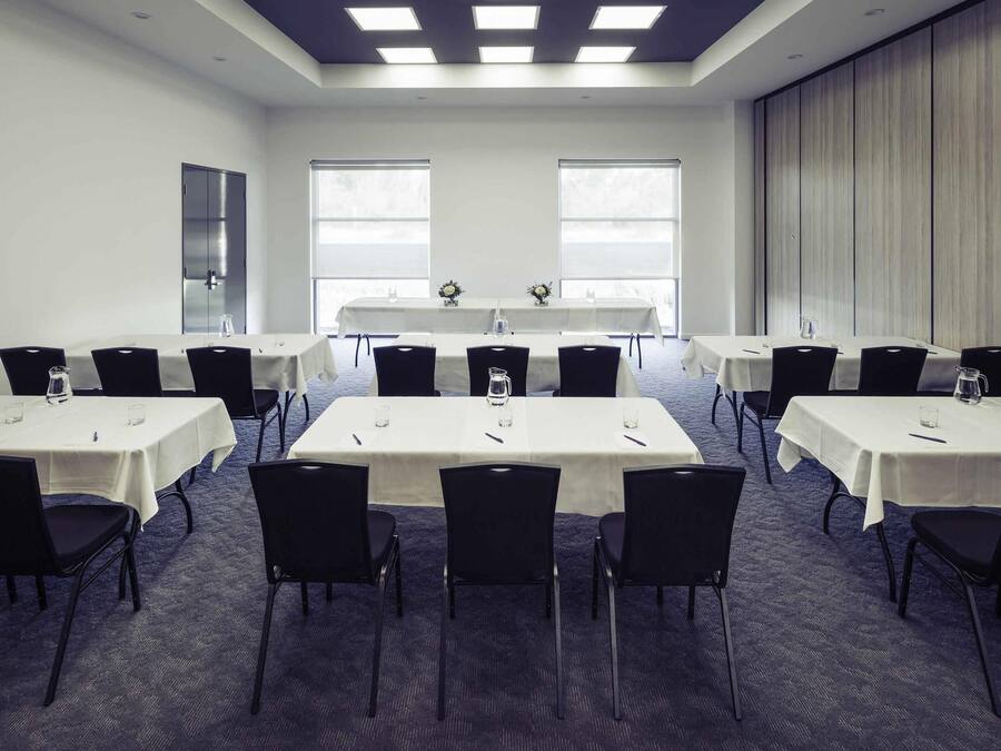 Sala de conferências com várias mesas e cadeiras, janelas amplas e design moderno no teto.
