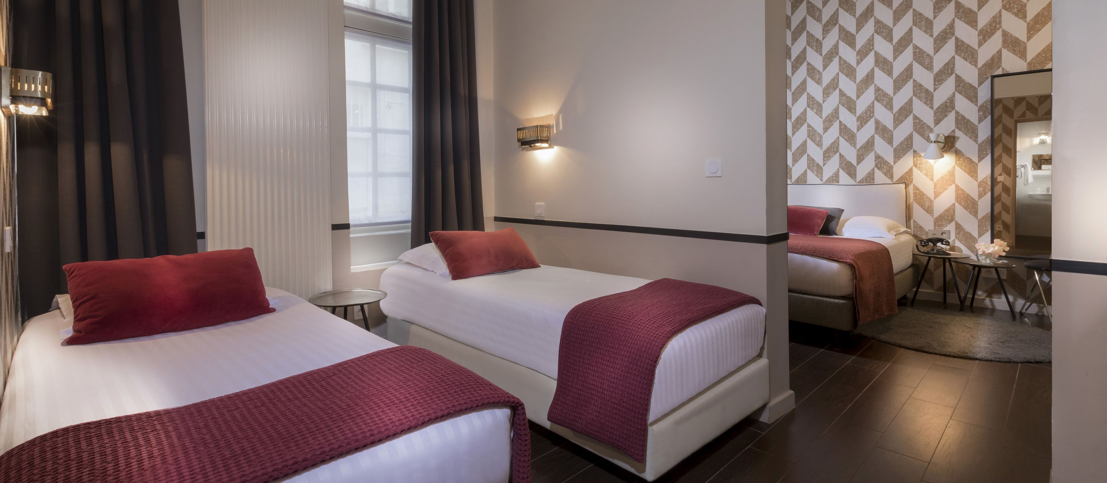 Quarto de hotel com duas camas, cada uma com almofadas e manta vermelhas, uma pequena mesa redonda, uma cadeira e um espelho.