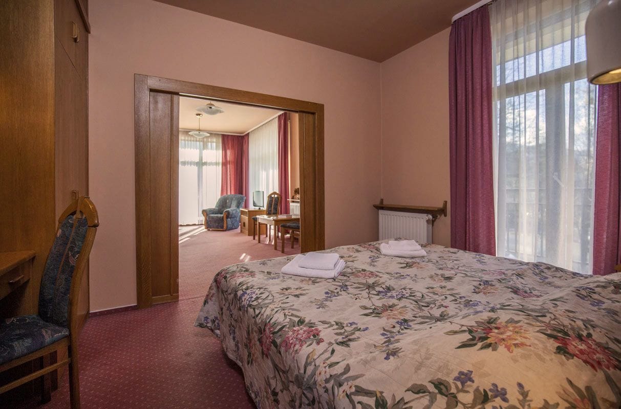 Quarto de hotel com uma cama grande, uma cadeira de madeira, uma escrivaninha e vista para outro quarto através de uma porta aberta.