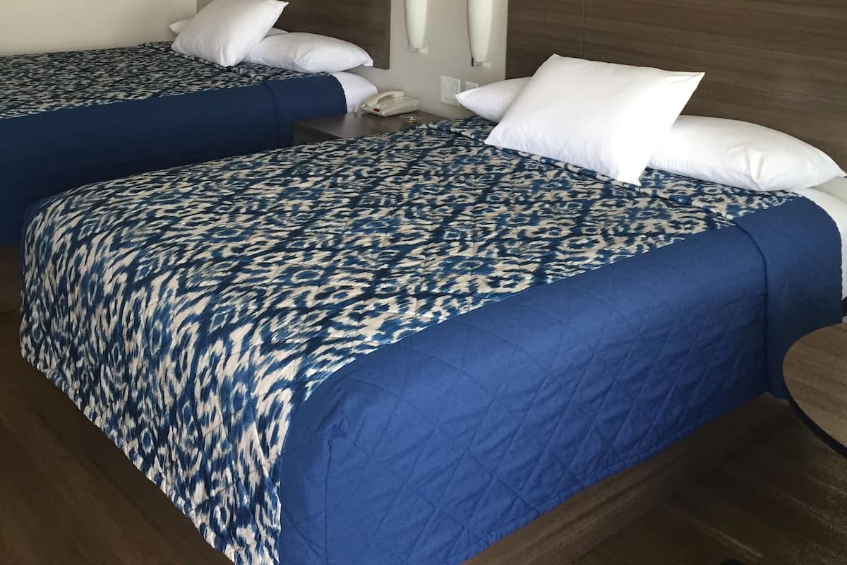 Quarto de hotel com duas camas, cada uma com roupa de cama estampada em azul e almofadas brancas. Há duas mesas de cabeceira, um telefone fixado na parede e um quadro na parede.