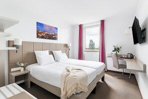 Quarto de hotel moderno com cama, mesa de cabeceira, escrivaninha, cadeira e televisão fixada na parede.