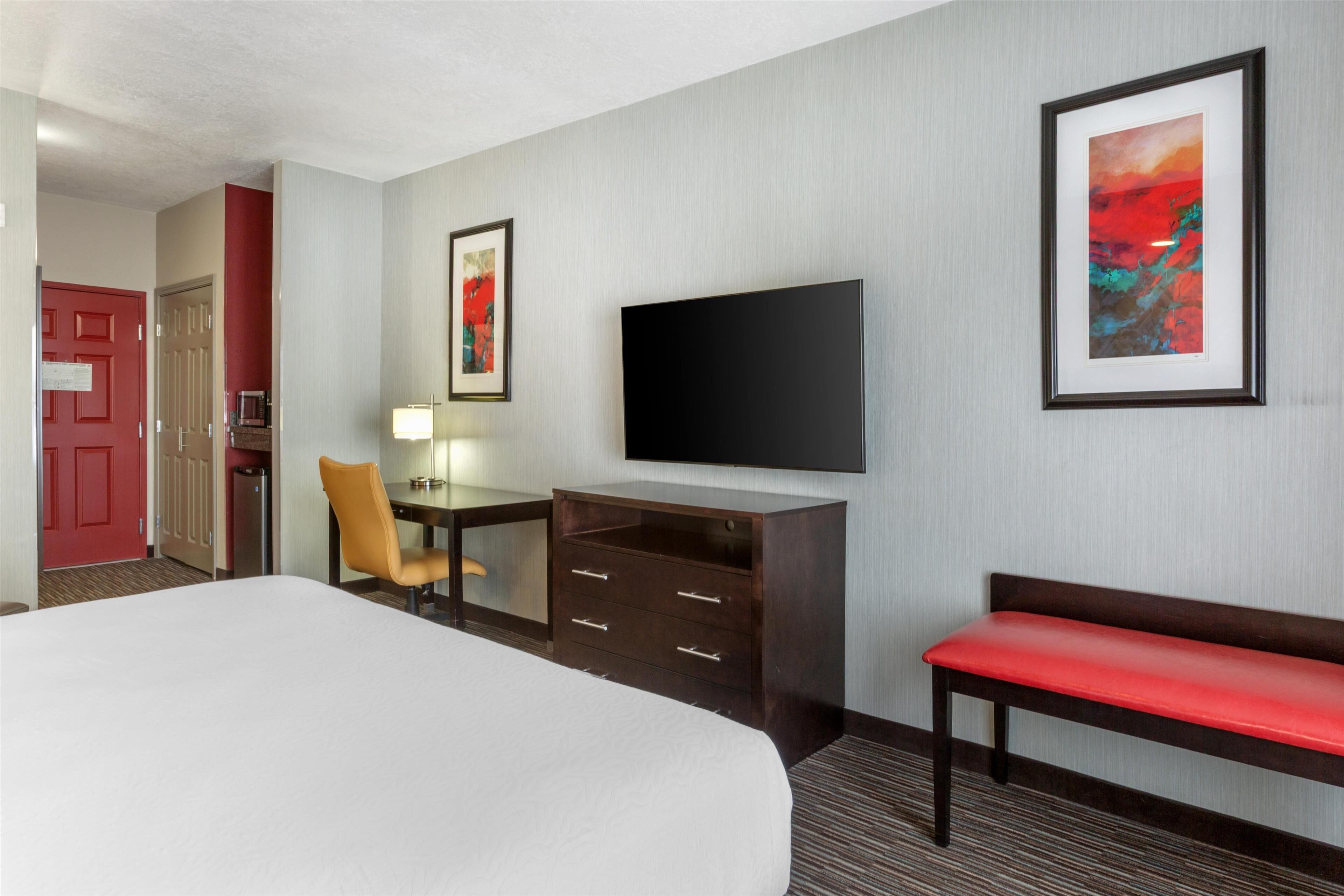 Quarto de hotel com uma cama grande, uma escrivaninha com cadeira, uma televisão e um banco.