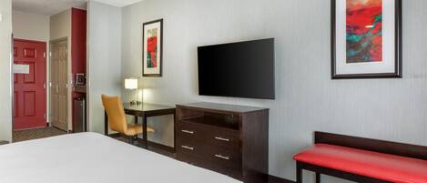 Quarto de hotel com uma cama grande, uma escrivaninha com cadeira, uma televisão e um banco.