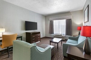 Quarto de hotel com televisão de tela plana, uma escrivaninha com luminária, duas cadeiras, um sofá, uma mesa de centro e uma janela com cortinas.