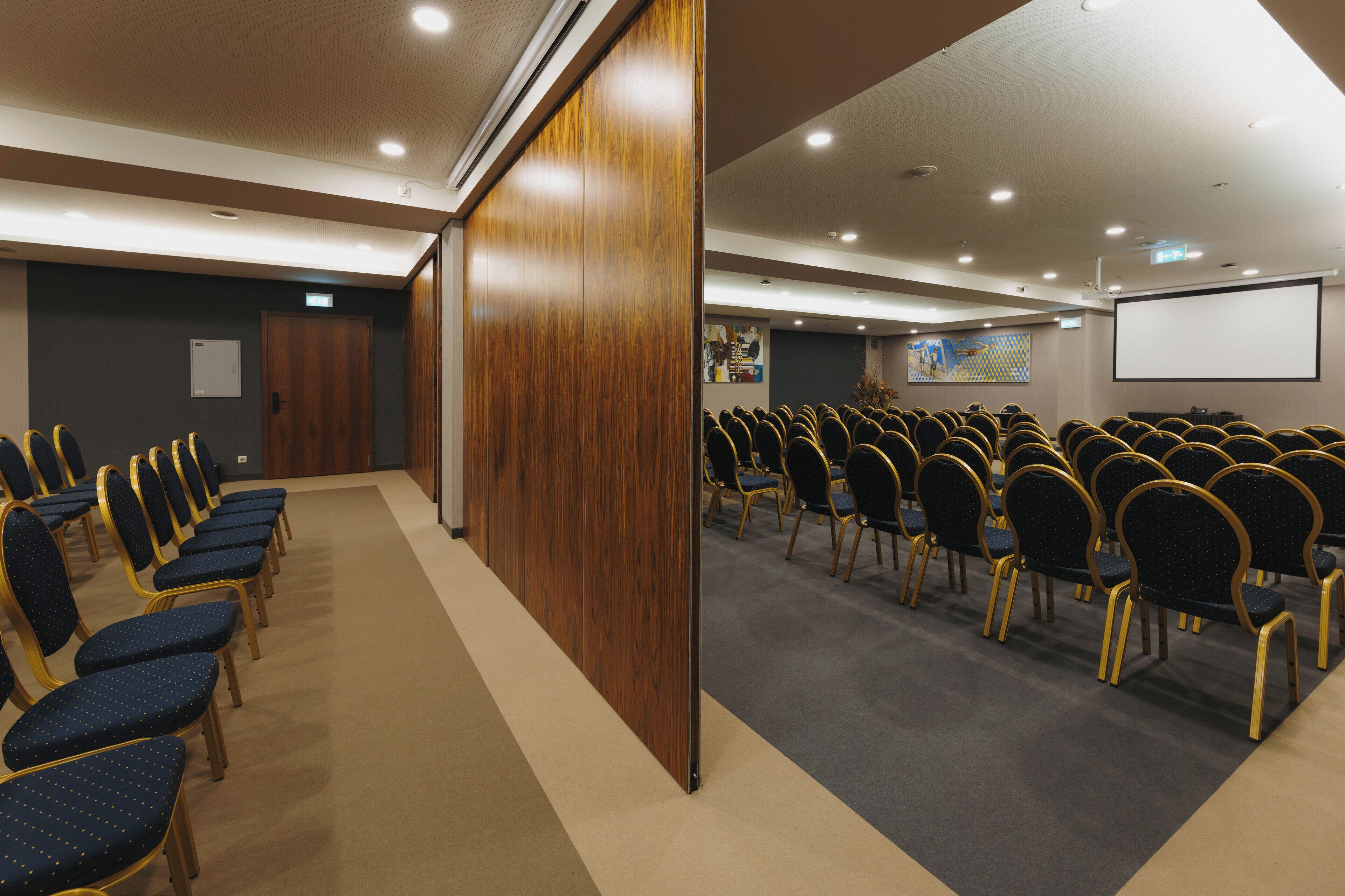Sala de conferências com fileiras de cadeiras, divisória de madeira e tela de projeção.