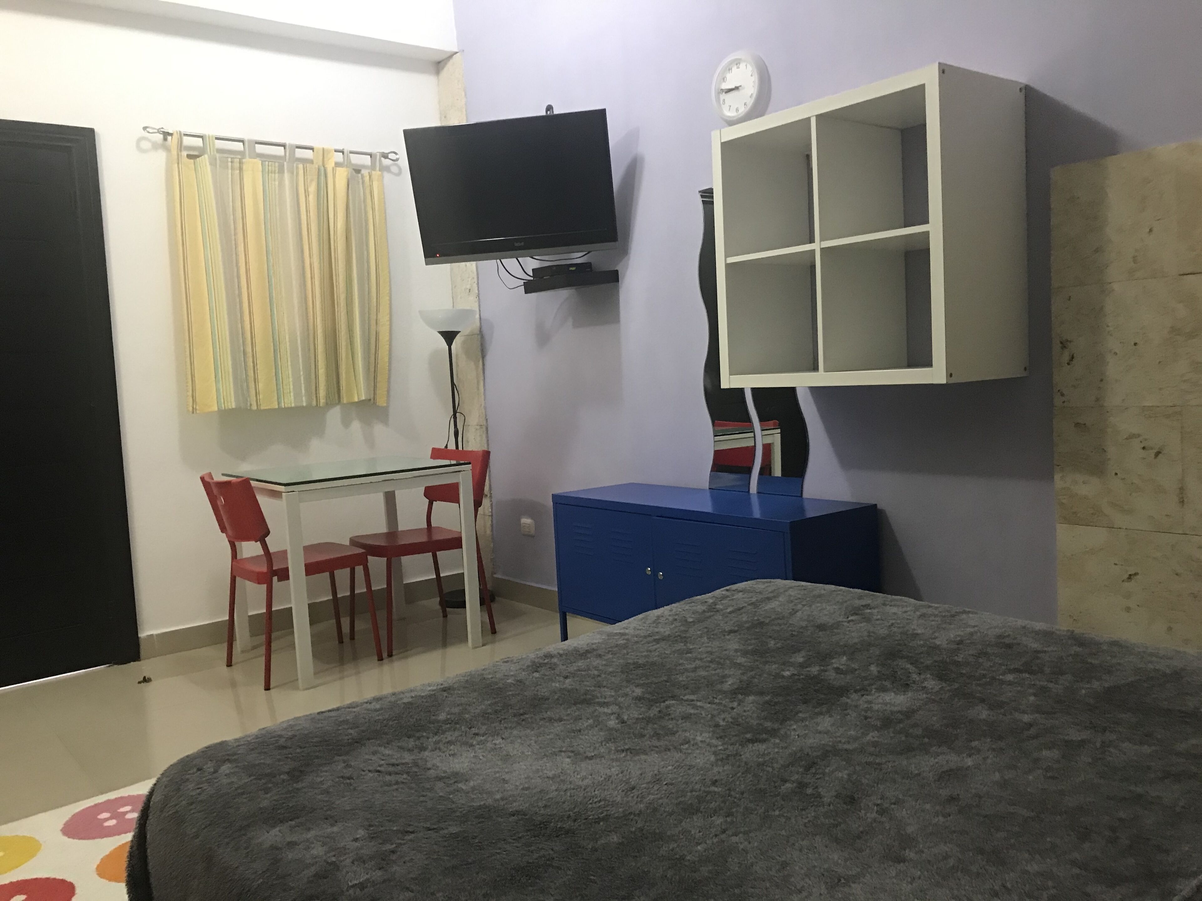 Quarto com uma cama, uma escrivaninha com duas cadeiras, uma televisão na parede e um armário.