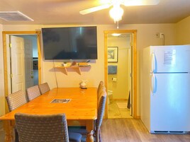 Uma sala de jantar com uma mesa de madeira, cadeiras, uma televisão montada acima e uma geladeira à direita.