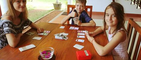 Três pessoas jogando um jogo de cartas em uma mesa de madeira, com uma tigela de flores e uma caixa vermelha em cima.