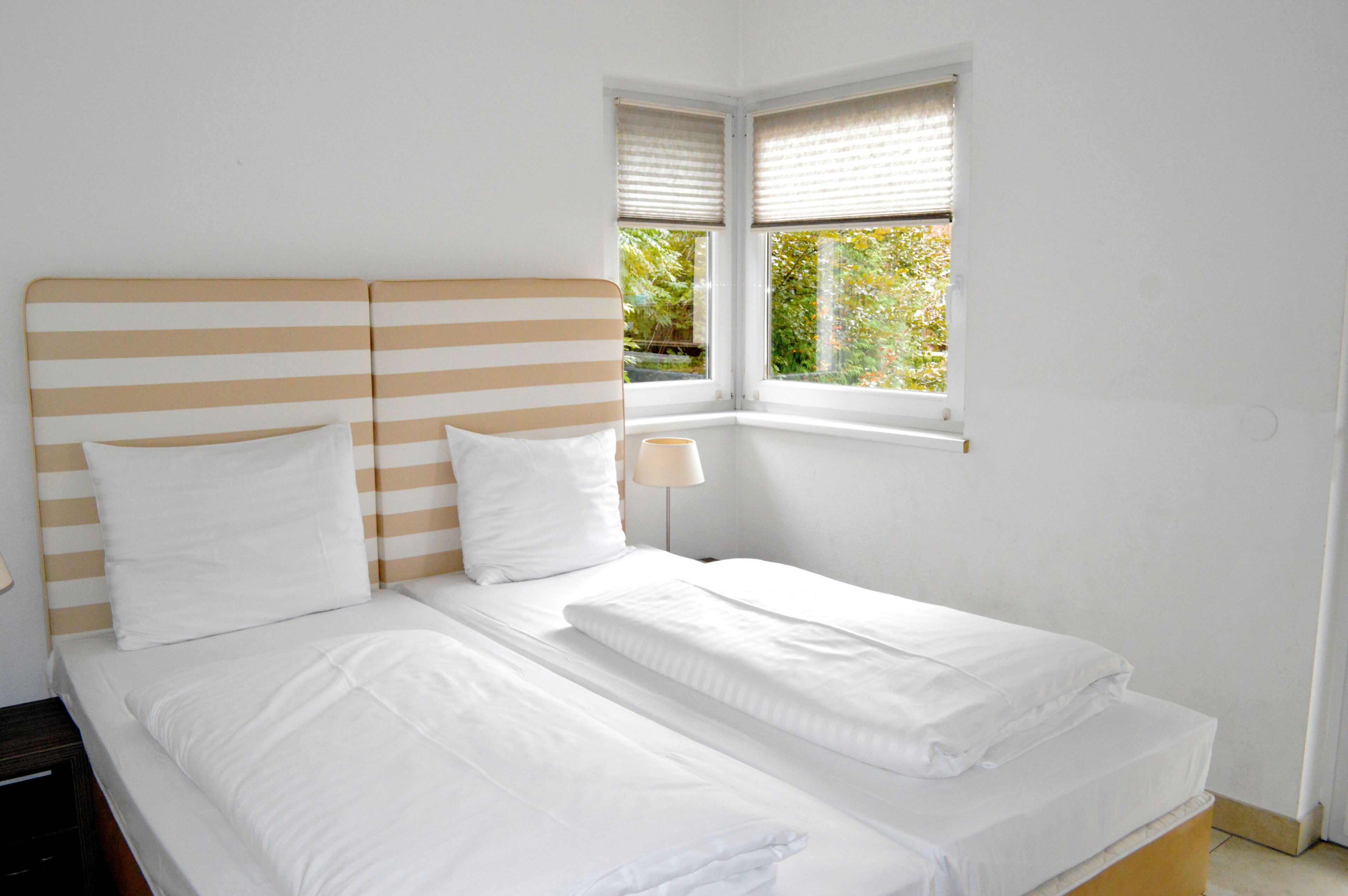 Cama de casal com cabeceira listrada, roupa de cama branca, mesa de cabeceira com abajur e janela com persianas.