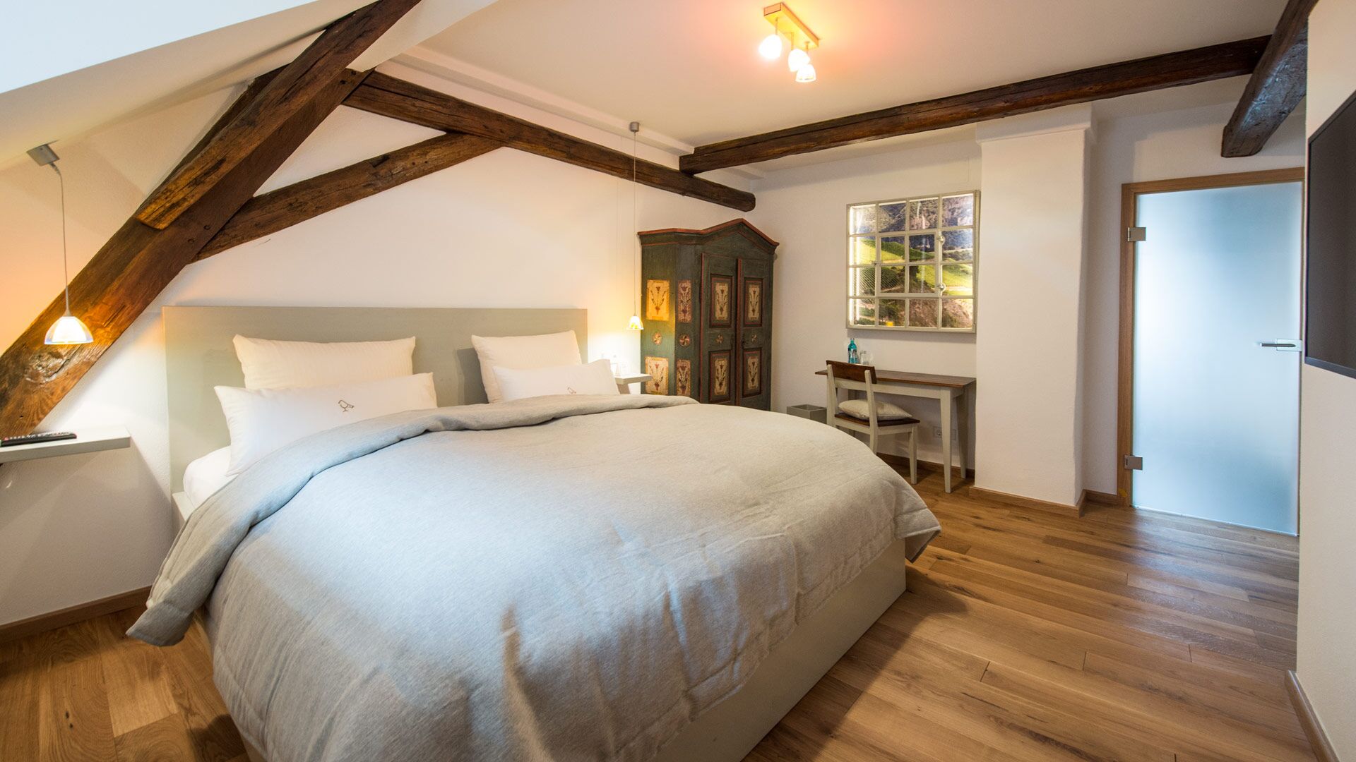 Quarto com uma cama grande, vigas de madeira, uma escrivaninha e uma janela com vista.