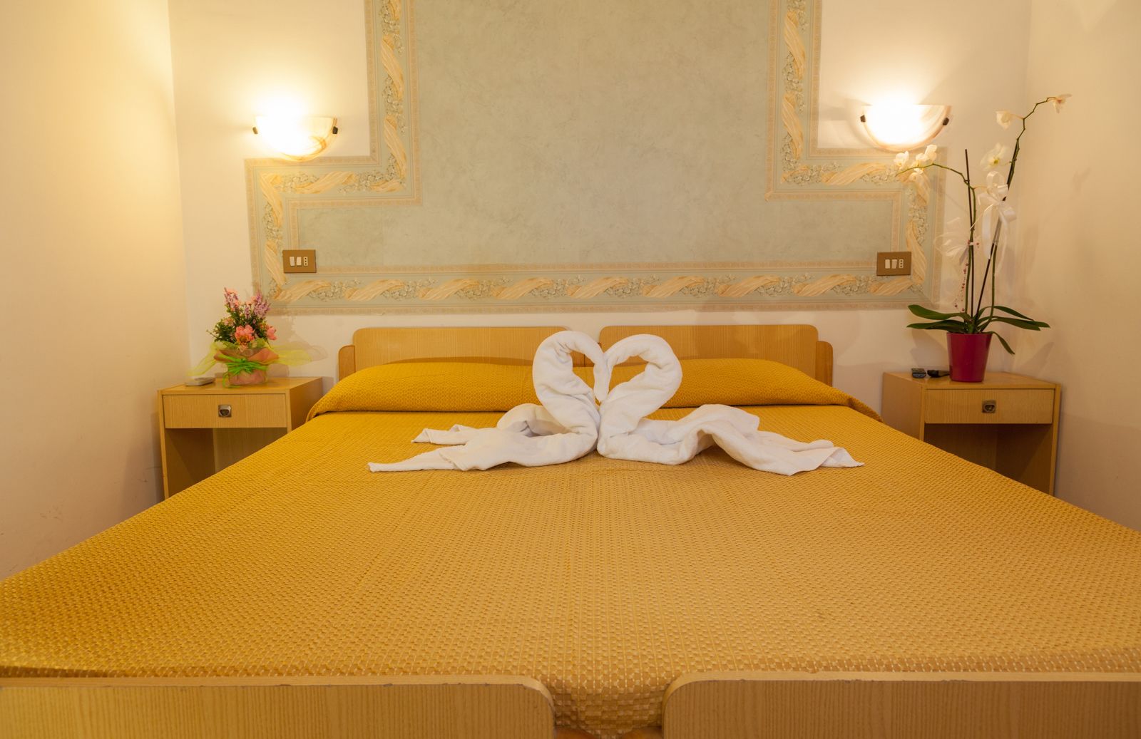 Quarto de hotel com uma cama, duas mesas de cabeceira, luminária fixada na parede e um vaso de planta.