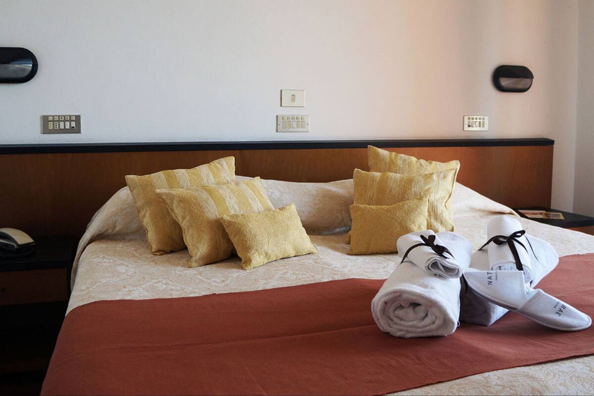Cama bem arrumada com almofadas, manta vermelha e toalhas enroladas em um quarto de hotel.