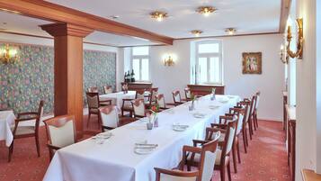 Uma sala de jantar com mesas compridas dispostas para refeição, cadeiras de madeira e um papel de parede estampado.