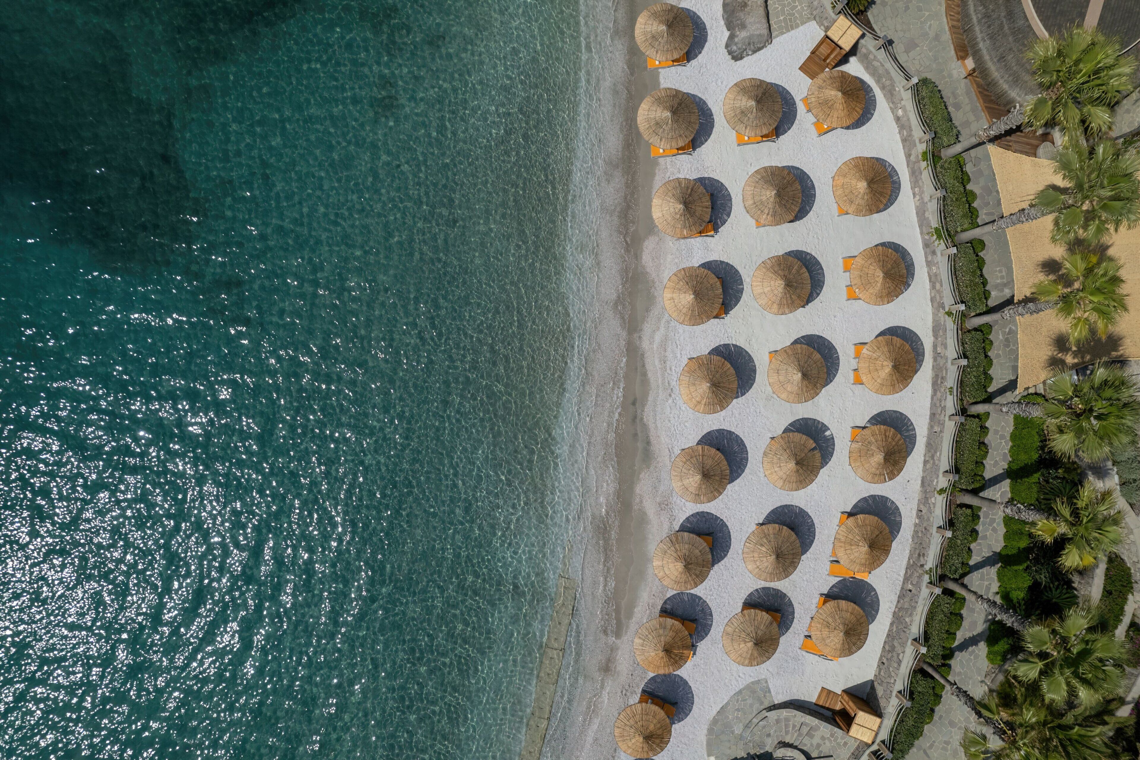 Vista aérea de uma praia com fileiras de guarda-sóis e espreguiçadeiras, um calçadão pavimentado e palmeiras.