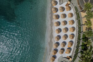 Vista aérea de uma praia com fileiras de guarda-sóis e espreguiçadeiras, um calçadão pavimentado e palmeiras.