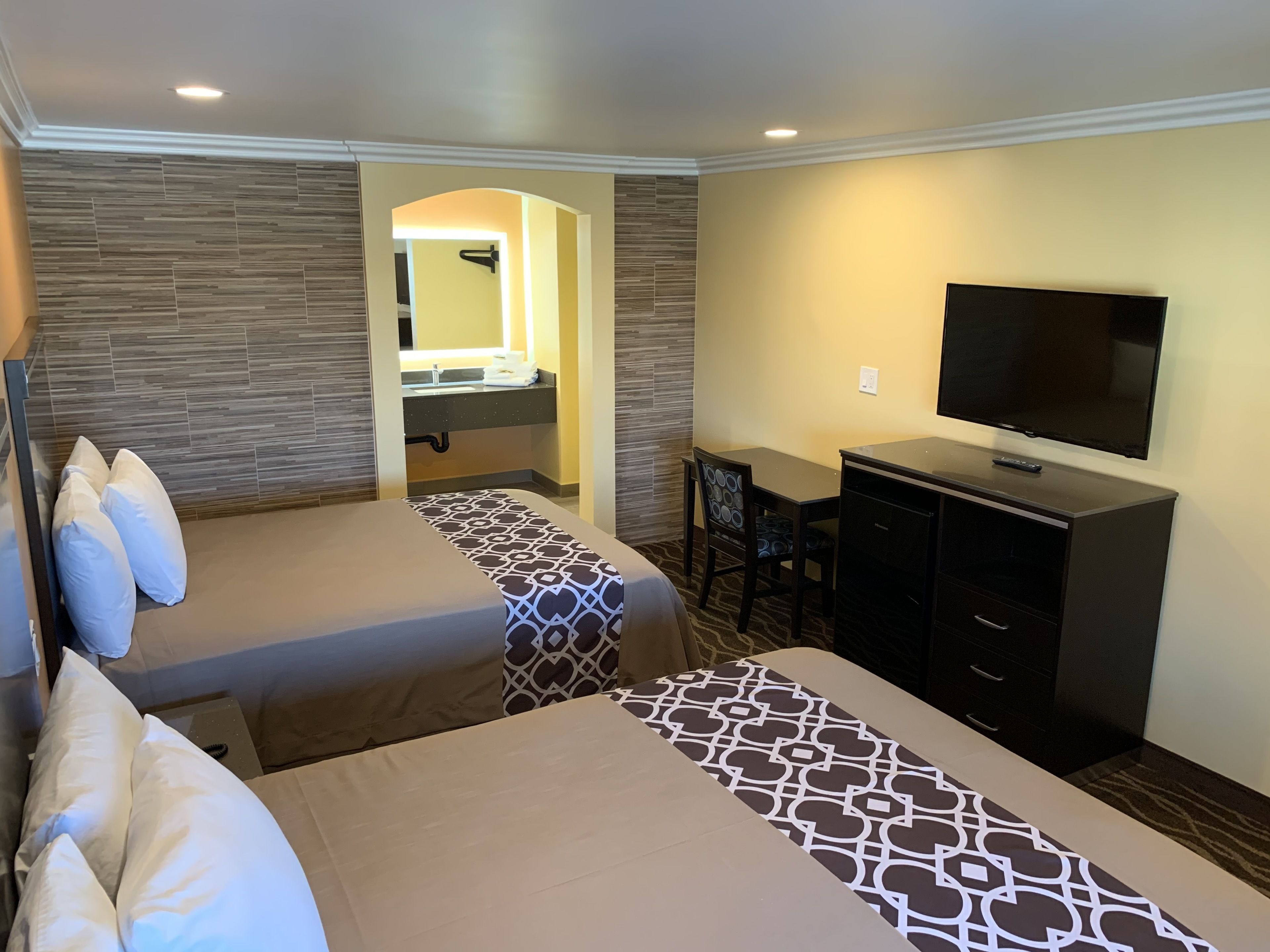 Quarto de hotel com duas camas, uma escrivaninha, televisão e banheiro visível por um arco.