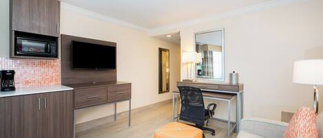 Quarto de hotel moderno com cozinha compacta, TV de tela plana, escrivaninha com cadeira e um espelho grande.