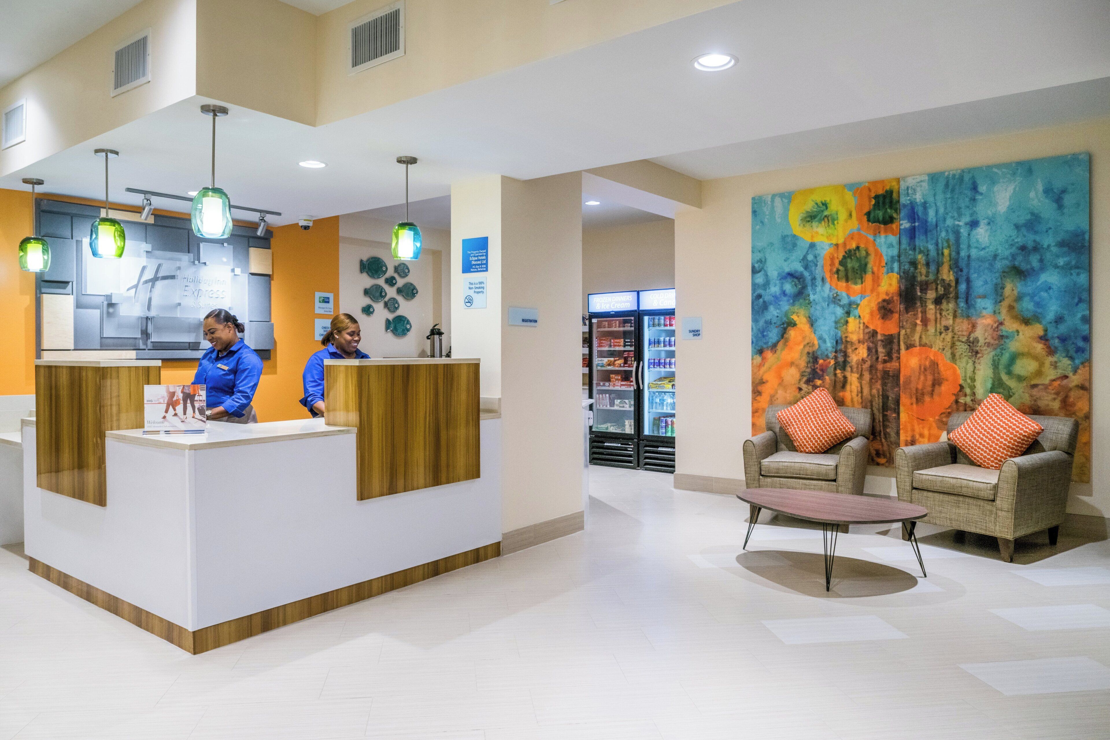 Área de recepção de hotel com balcão, dois funcionários, máquina de café, refrigerador e um quadro na parede.