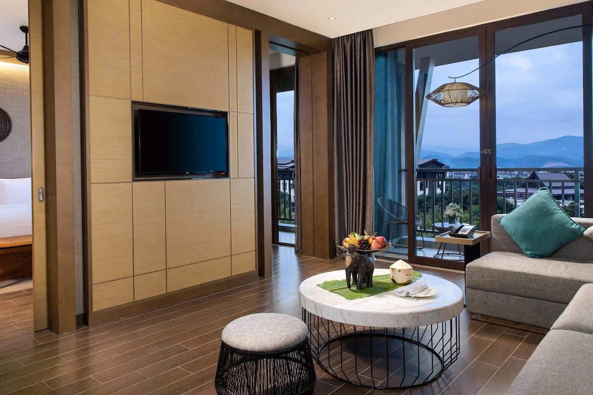 Quarto de hotel moderno com televisão de tela plana, uma mesa de centro redonda com uma estatueta de elefante decorativa, um sofá com almofada verde e uma varanda com vista para as montanhas.