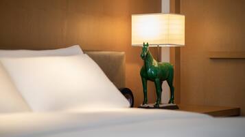 Uma cama com lençóis brancos, uma luminária em formato de cavalo verde e um relógio fixado na parede.