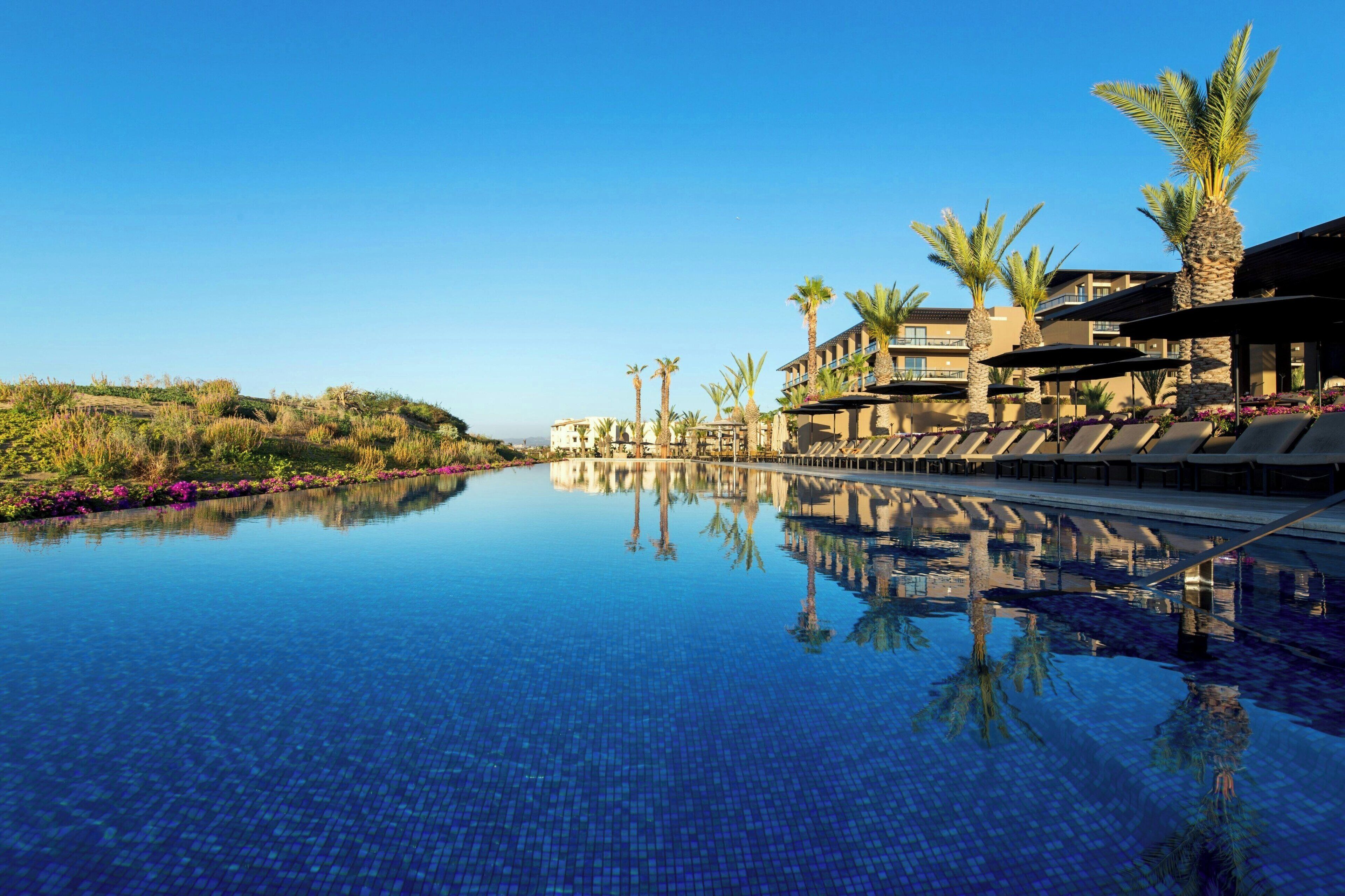 Uma piscina infinita azul límpida com espreguiçadeiras e palmeiras, refletindo os edifícios e o céu ao redor.