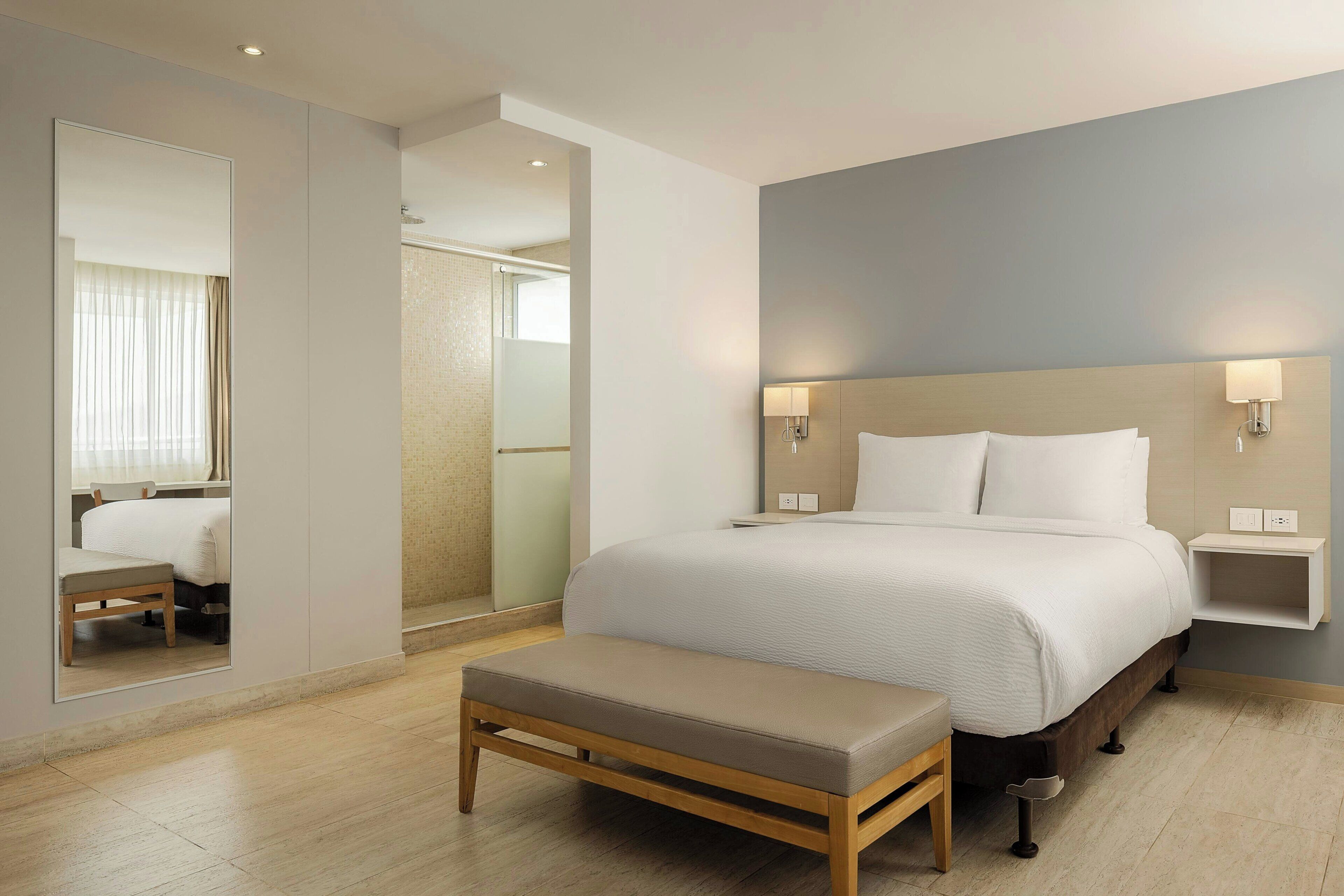 Quarto de hotel moderno com cama grande, mesinhas de cabeceira, um banco, espelho e área de chuveiro.