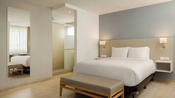 Quarto de hotel moderno com cama grande, mesinhas de cabeceira, um banco, espelho e área de chuveiro.
