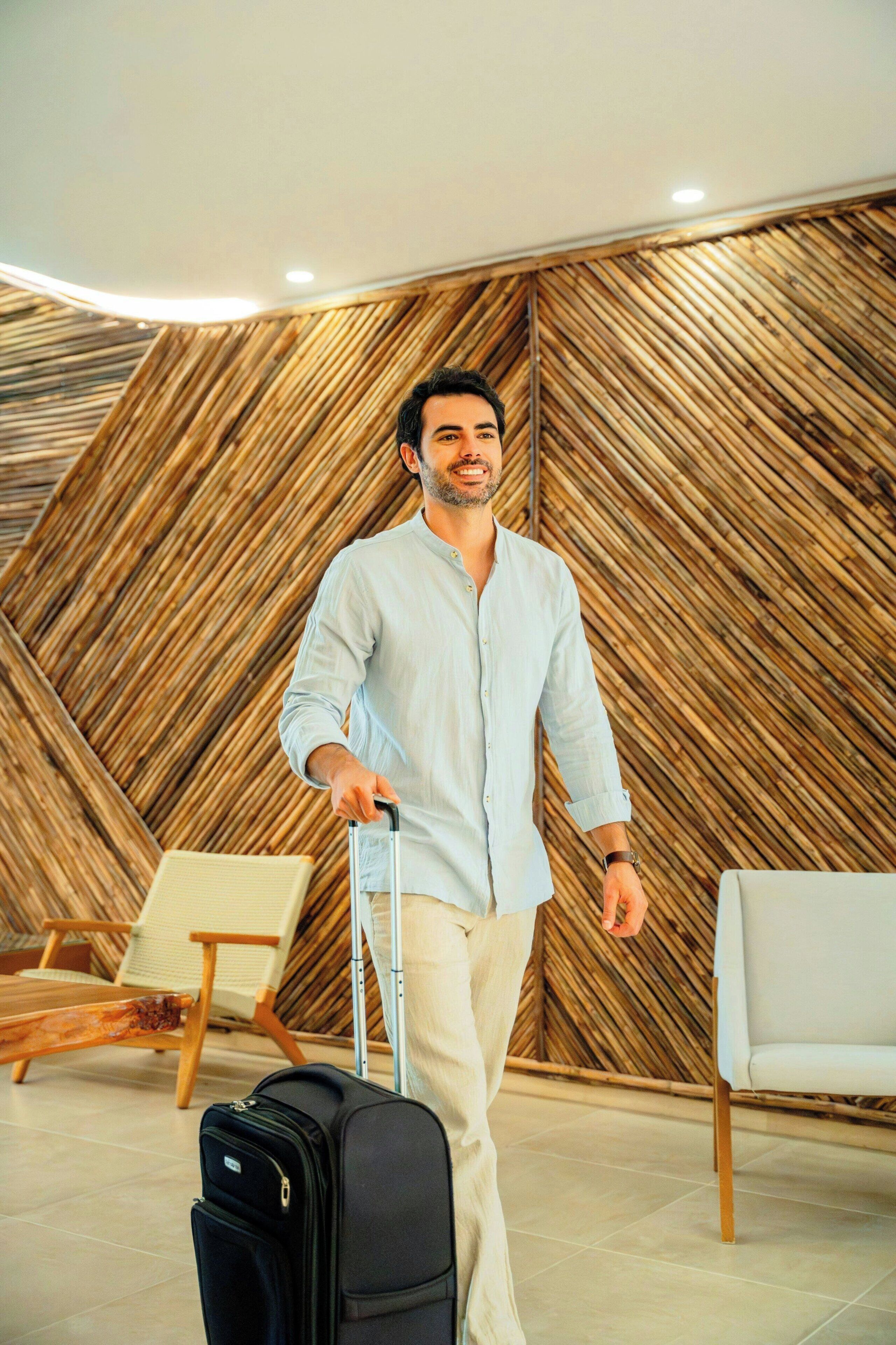 Um homem com uma mala em um quarto com parede de padrão de bambu e duas cadeiras brancas.